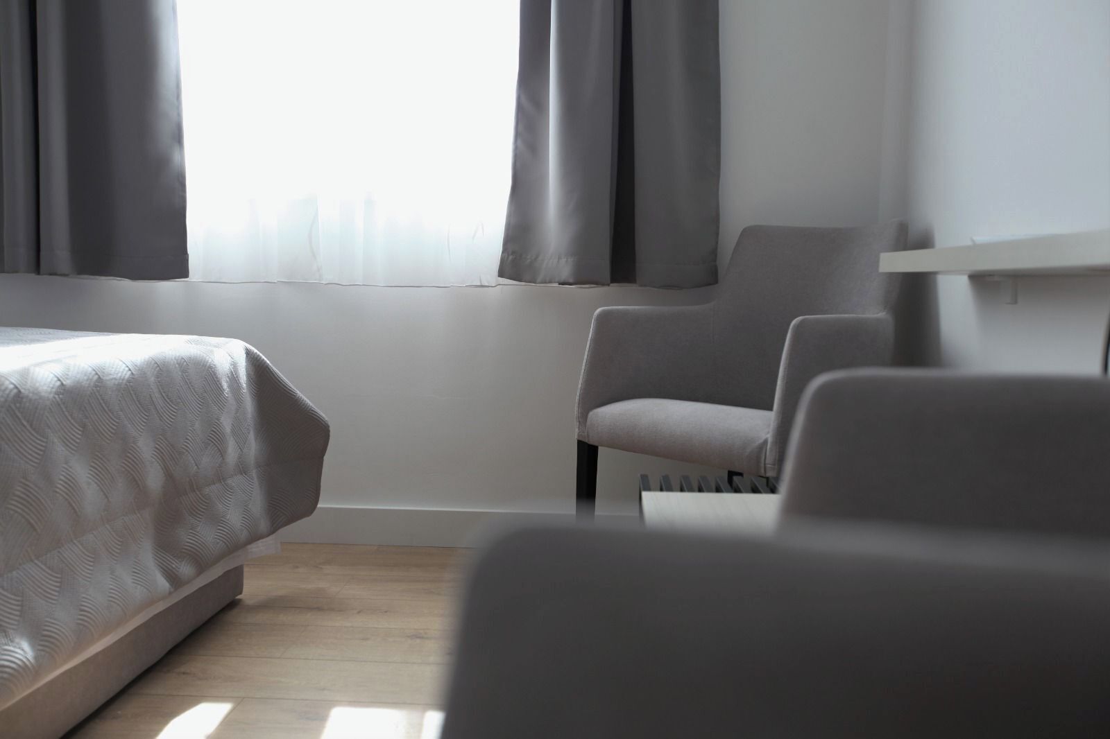 Quarto de hotel moderno com uma cama, dois poltronas, uma mesa pequena e uma janela com cortinas.