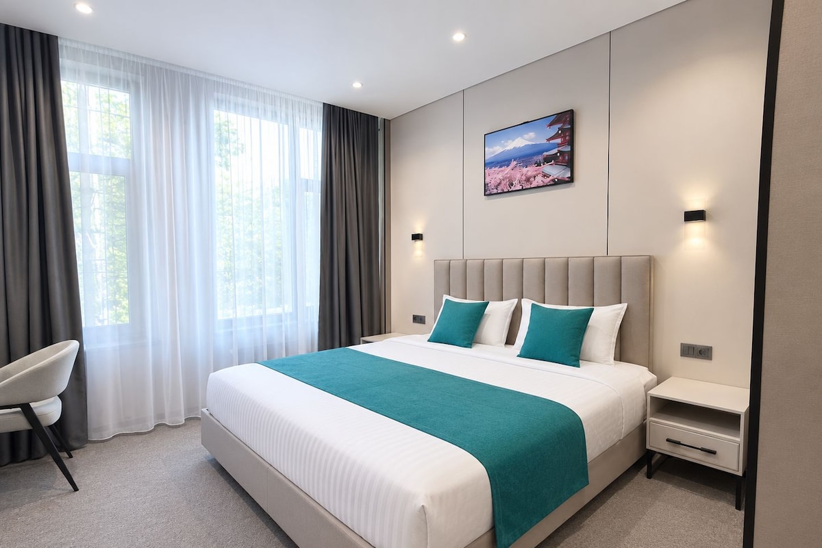 Quarto de hotel com uma cama grande, uma mesa de trabalho e uma cadeira. Há um quadro na parede e uma janela com cortinas.