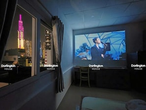 Um quarto com uma tela grande exibindo uma paisagem urbana e um artista, uma janela com vista para um edifício alto, e o texto 'Darlington HOUSE' na tela.