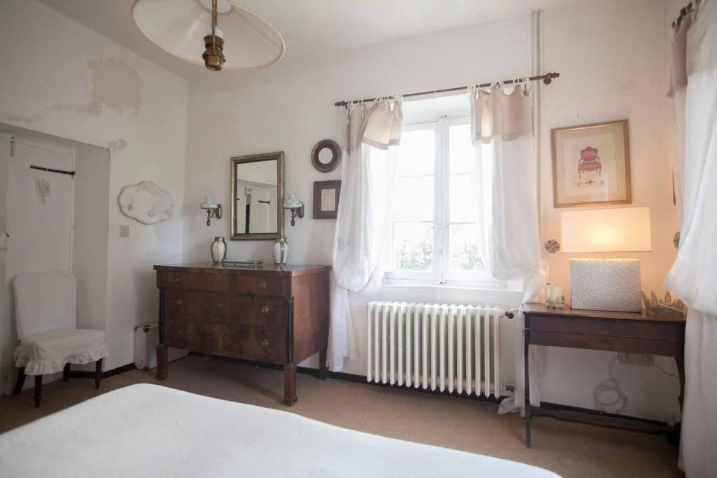 Quarto com cama, penteadeira, espelho, janela com cortinas, radiador e mesa de cabeceira com abajur.