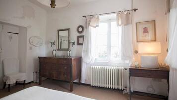 Quarto com cama, penteadeira, espelho, janela com cortinas, radiador e mesa de cabeceira com abajur.
