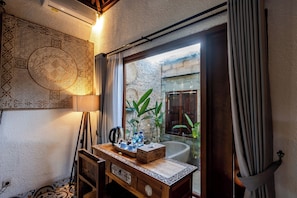 Um banheiro com banheira, um espelho com bancada de madeira e bandeja de amenities, parede com padrão decorativo e vista para área externa com plantas.