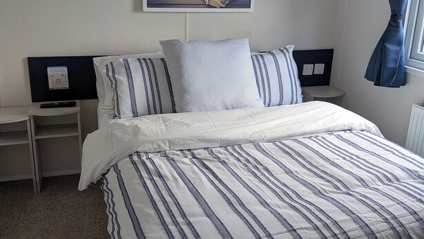 Cama bem arrumada com roupa de cama listrada, mesa de cabeceira com controle remoto e uma obra de arte na parede.