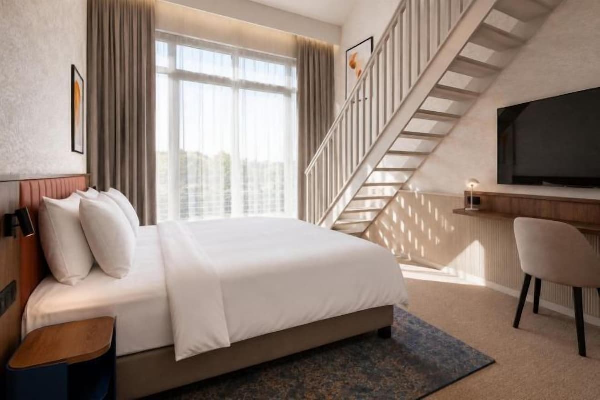 Quarto de hotel com cama, escrivaninha, cadeira, televisão e escada.