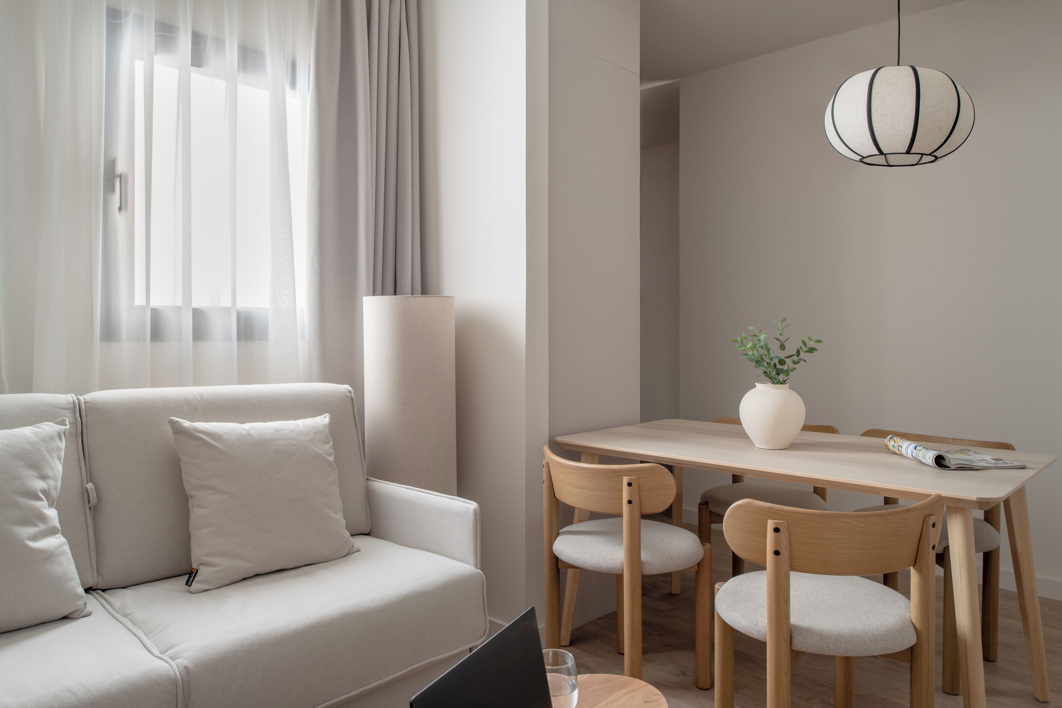 Uma sala de estar moderna com um sofá, uma mesa de jantar com cadeiras, um abajur e um vaso com planta.