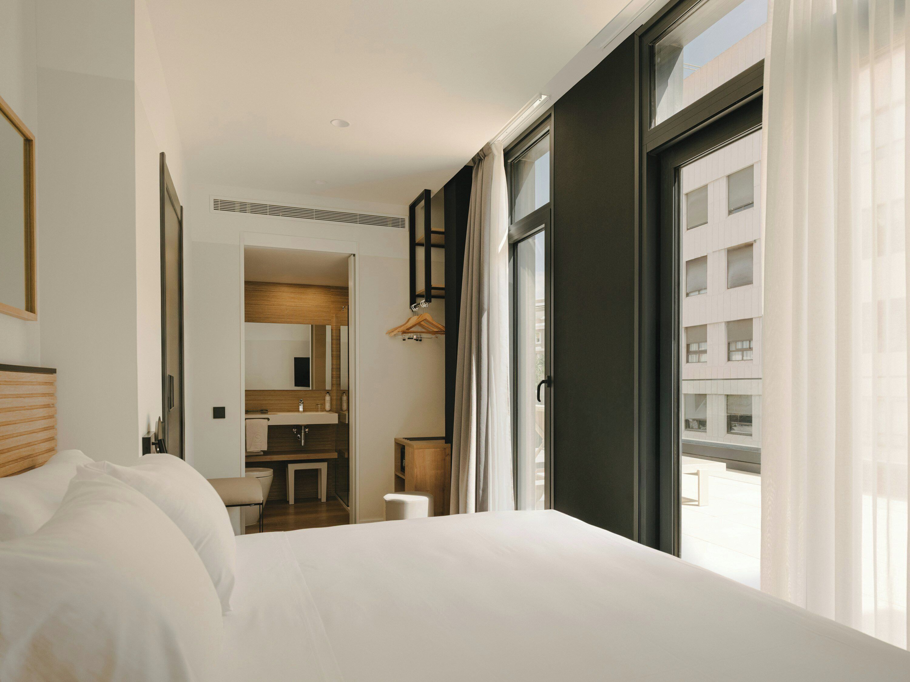 Quarto de hotel com uma cama grande, banheiro com espelho e pia, e vista para edifícios pela janela.
