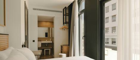 Quarto de hotel com uma cama grande, banheiro com espelho e pia, e vista para edifícios pela janela.