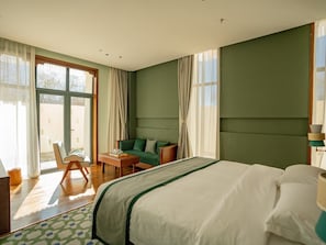 Quarto de hotel com uma cama grande, um sofá, uma mesinha e uma cadeira. Há uma janela com cortinas, e a parede do quarto é verde.