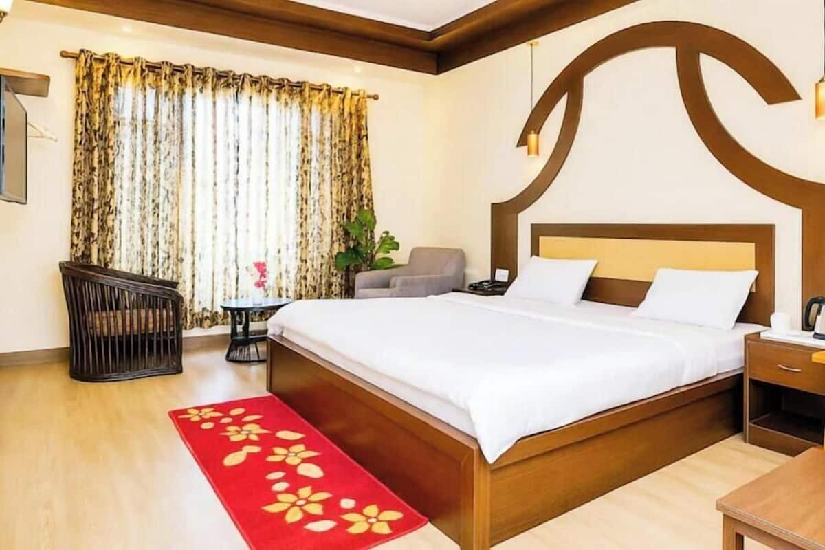 Quarto de hotel com uma cama grande, cabeceira de madeira, tapete floral vermelho, uma pequena mesa com um vaso e uma cadeira.