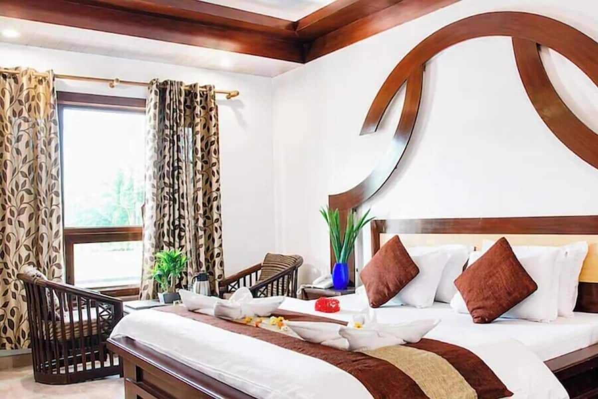 Quarto de hotel com uma cama grande, cabeceira de madeira, janela com cortinas, mesa de cabeceira e apoio para objetos pessoais.