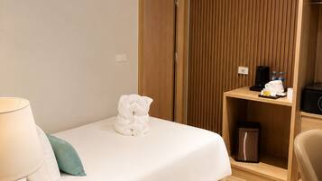 Quarto de hotel com cama, mesa de cabeceira com abajur, cadeira, guarda-roupa de madeira e prateleira pequena com produtos de higiene pessoal.