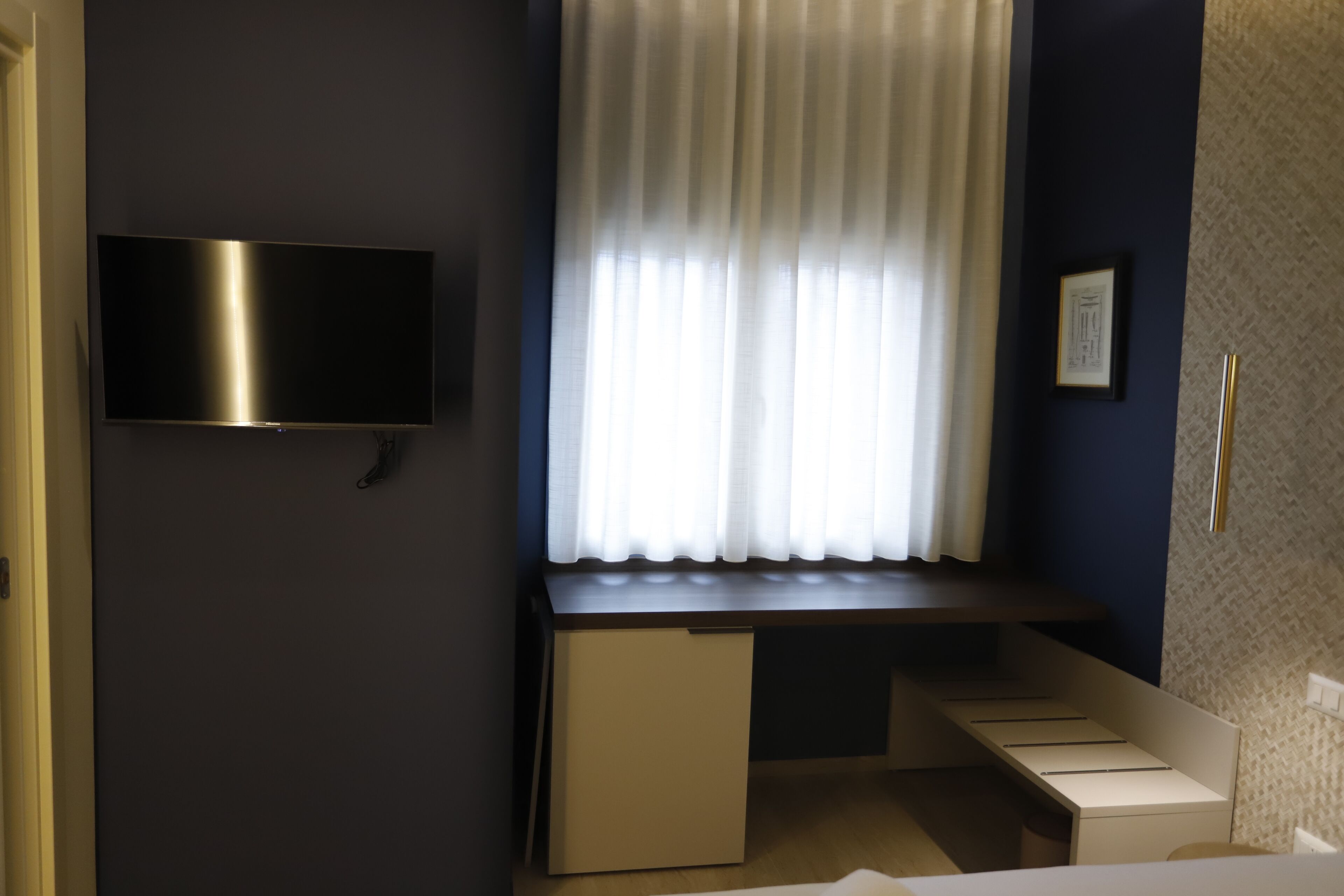 Quarto com televisão de tela plana na parede, janela com cortinas leves, escrivaninha embutida com cadeira e parede com padrão.