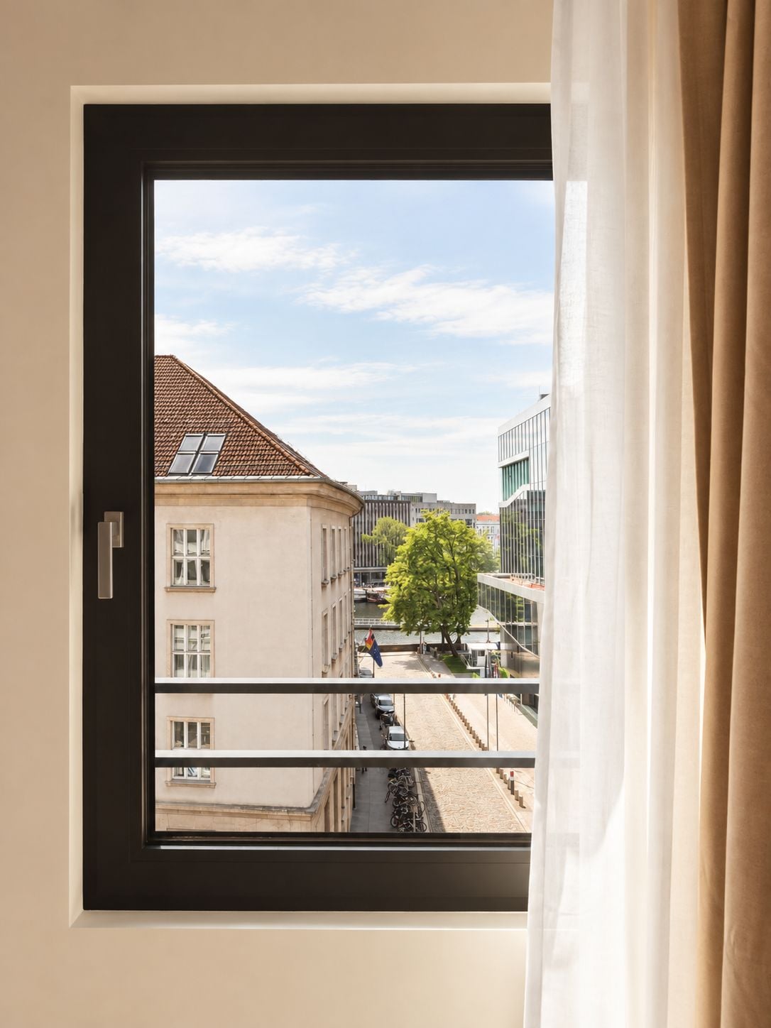 Janela com vista para um edifício com telhado de azulejos e uma árvore ao primeiro plano.