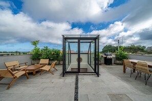 Terraço no telhado com estrutura de vidro, mobiliário de vime e churrasqueira.