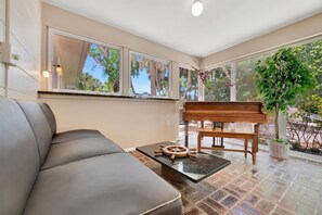 Uma sala de estar com sofá, piano, mesa de centro com um leme e uma grande janela com vista para palmeiras.