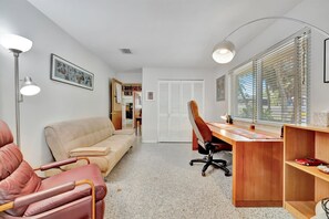 Quarto com sofá bege, secretária de madeira, cadeira de escritório vermelha, luminária de chão e janela com persianas.