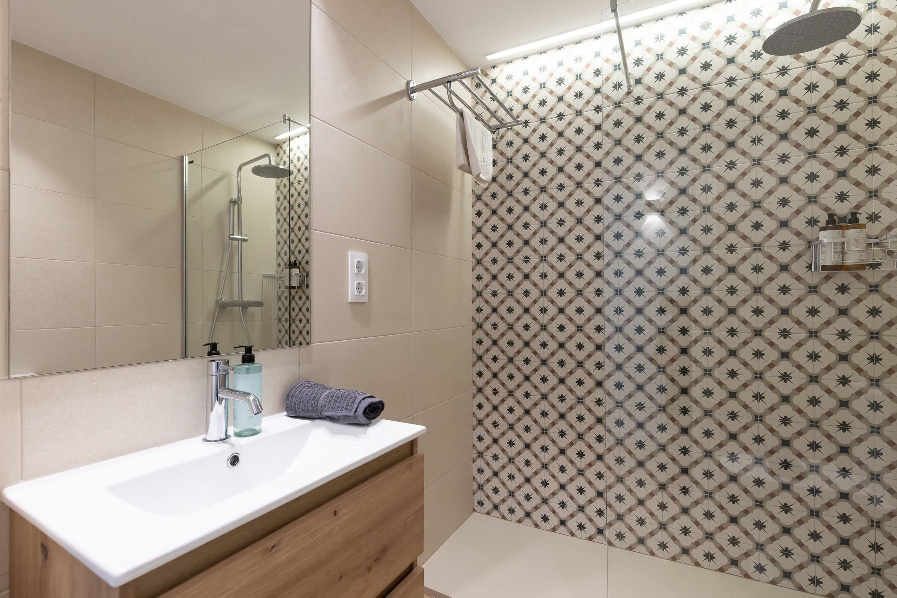 Casa de banho com lavatório branco, espelho, chuveiro com chuveiro de efeito chuva e azulejos estampados.