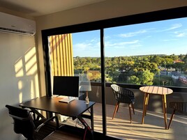 Quarto moderno com uma janela ampla, uma escrivaninha com computador, uma cadeira e uma pequena mesa.