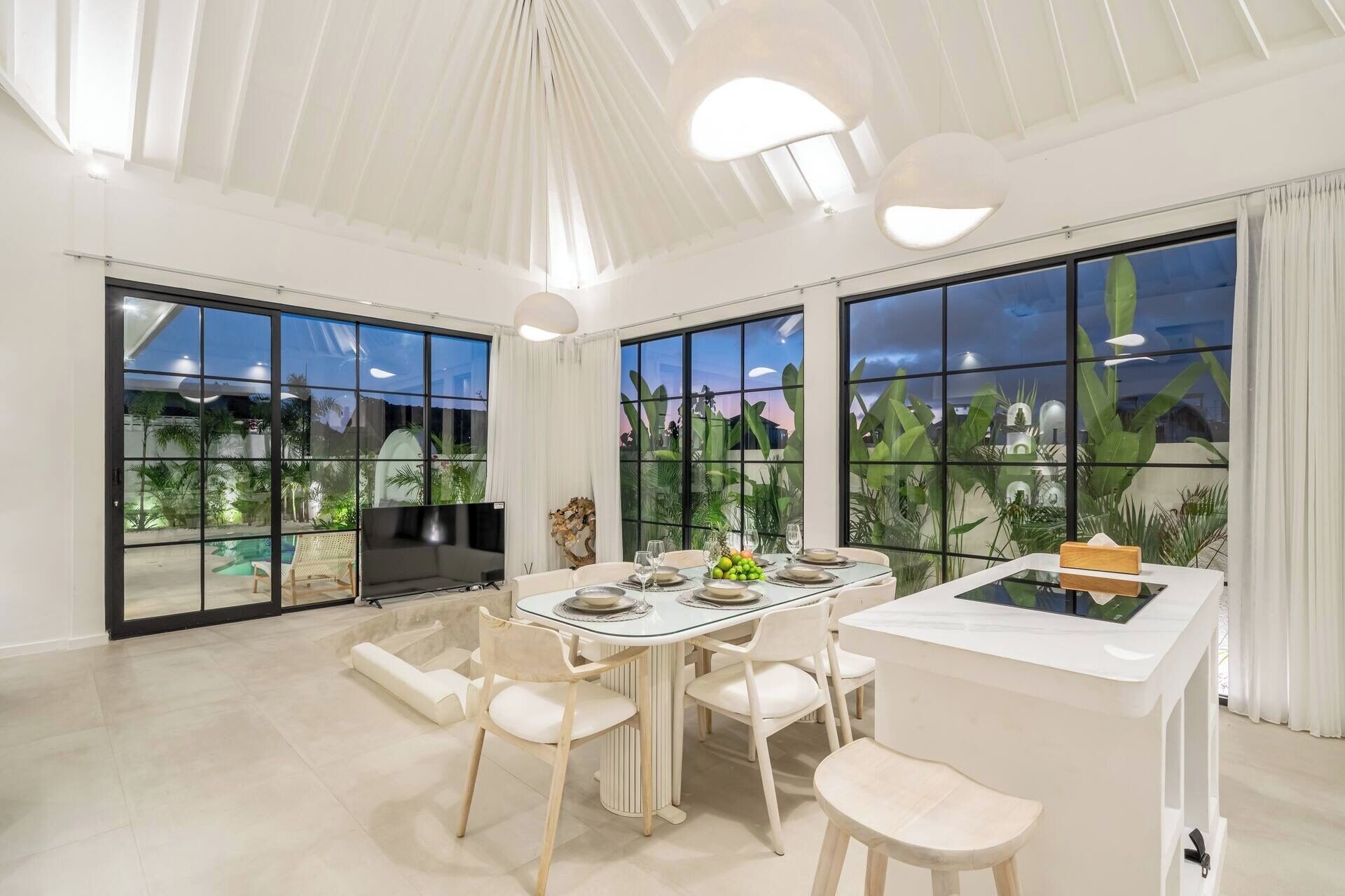 Uma sala de jantar espaçosa com uma mesa grande, cadeiras e uma ilha de cozinha, com grandes janelas que oferecem vista para uma piscina e vegetação.