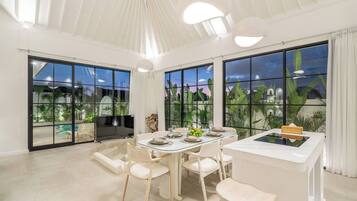 Uma sala de jantar espaçosa com uma mesa grande, cadeiras e uma ilha de cozinha, com grandes janelas que oferecem vista para uma piscina e vegetação.