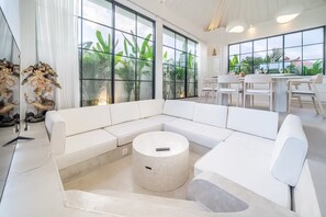 Uma sala de estar moderna com um sofá seccional branco, uma mesa de centro redonda e grandes janelas com vista para o verde.