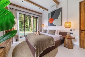 Quarto com uma cama grande, uma mesa redonda de madeira, um quadro e vista para árvores pela janela.
