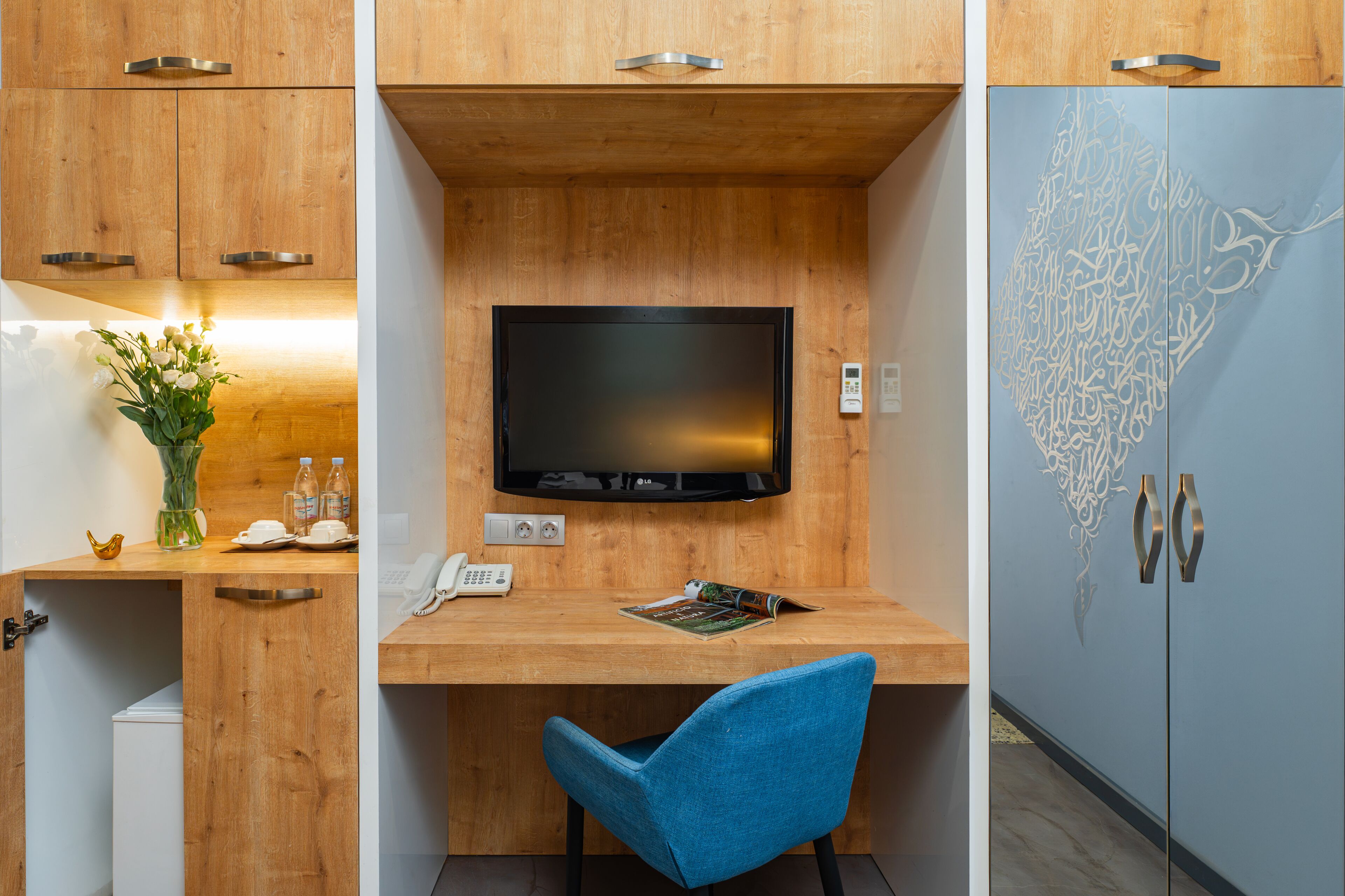 Quarto compacto com uma escrivaninha de madeira, uma cadeira azul, uma televisão e um frigorífico com decoração em relevo.