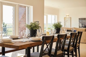 Uma sala de jantar com mesa de madeira posta para seis, plantas em vasos e vista para o exterior.