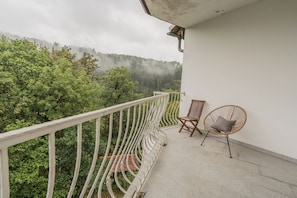 Uma varanda com guarda-corrimão branco, duas cadeiras de vime e vista para uma encosta arborizada.