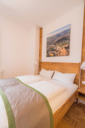 Quarto com cabeceira de madeira, cama grande com roupa de cama branca, detalhe verde no edredom, pintura pendurada na parede representando uma paisagem e mesa de cabeceira com abajur.