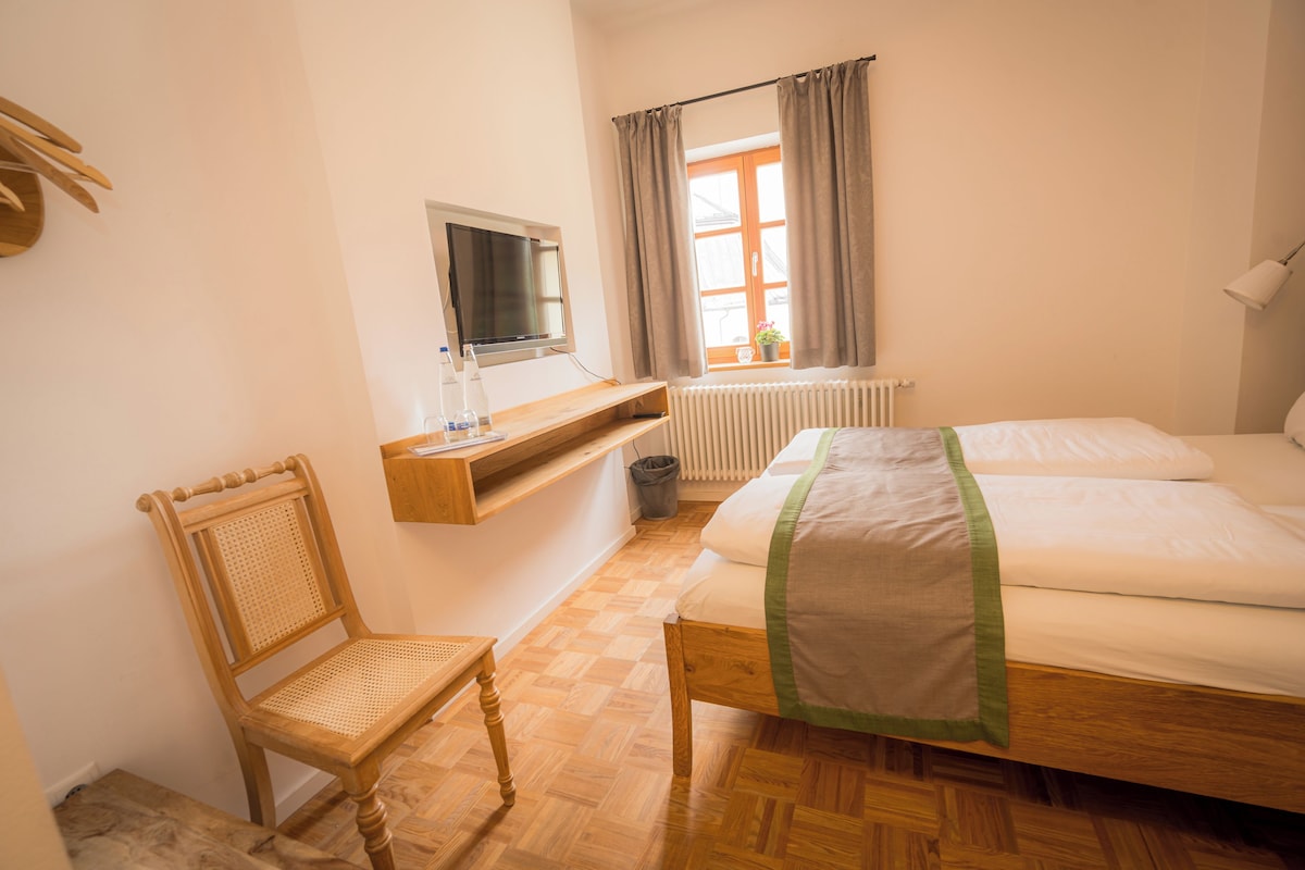 Quarto de hotel com chão de madeira, cama com colcha verde e branca, cadeira de madeira, televisão presa à parede e janela com cortinas.