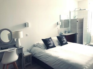 Quarto de hotel com cama, escrivaninha com luminária, cadeira, espelho e banheiro com chuveiro e vaso sanitário.