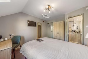 Quarto de hotel com cama, escrivaninha com cadeira, televisão, espelho, armário e chuveiro.
