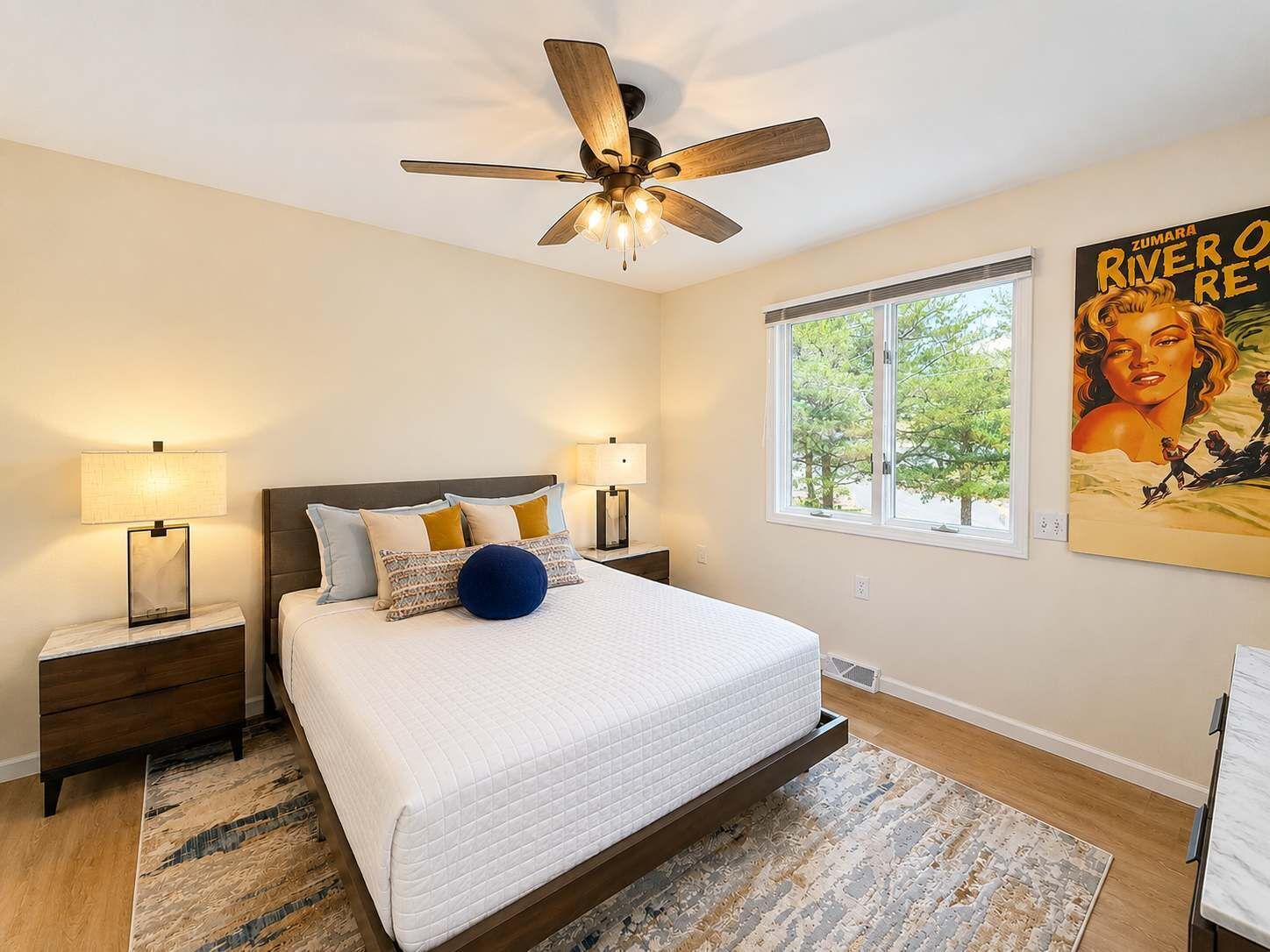 Quarto com cama, mesas de cabeceira, ventoinha no teto, janela e obras de arte na parede.