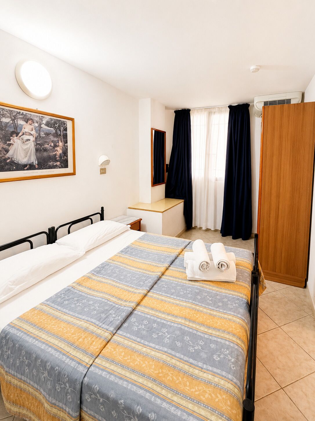 Quarto de hotel com cama, mesa de cabeceira, espelho, guarda-roupa e um quadro na parede.