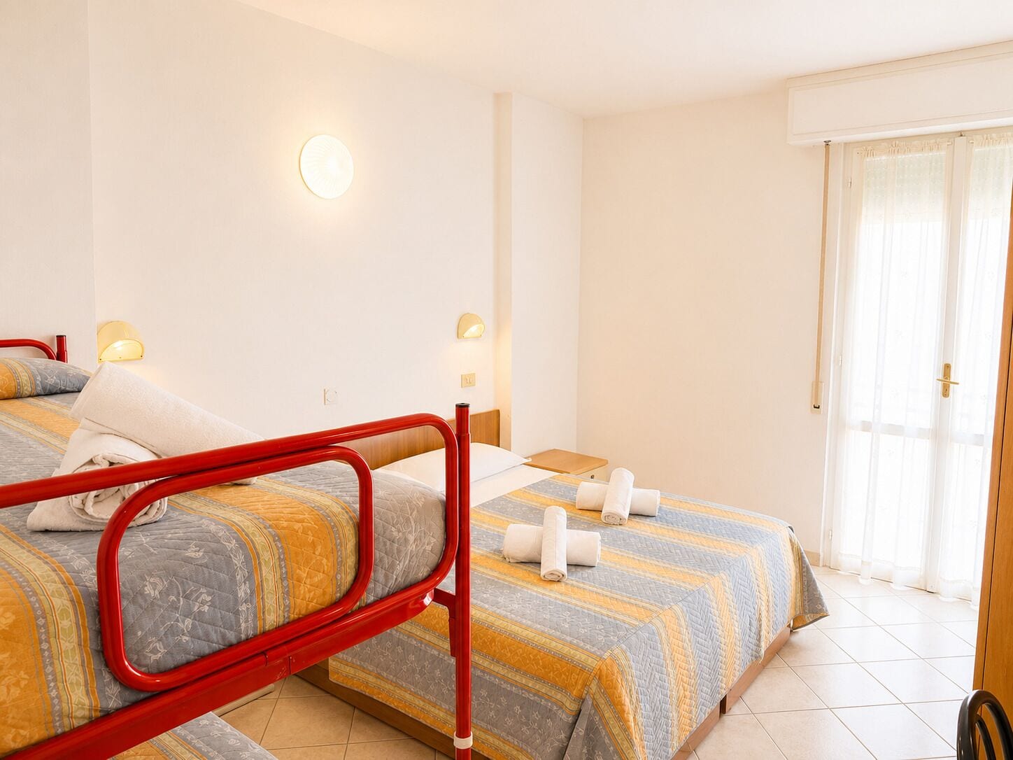 Quarto duplo com duas camas, estrutura de cama de metal vermelha, duas mesas de cabeceira, luminária fixada na parede e porta de vidro deslizante.