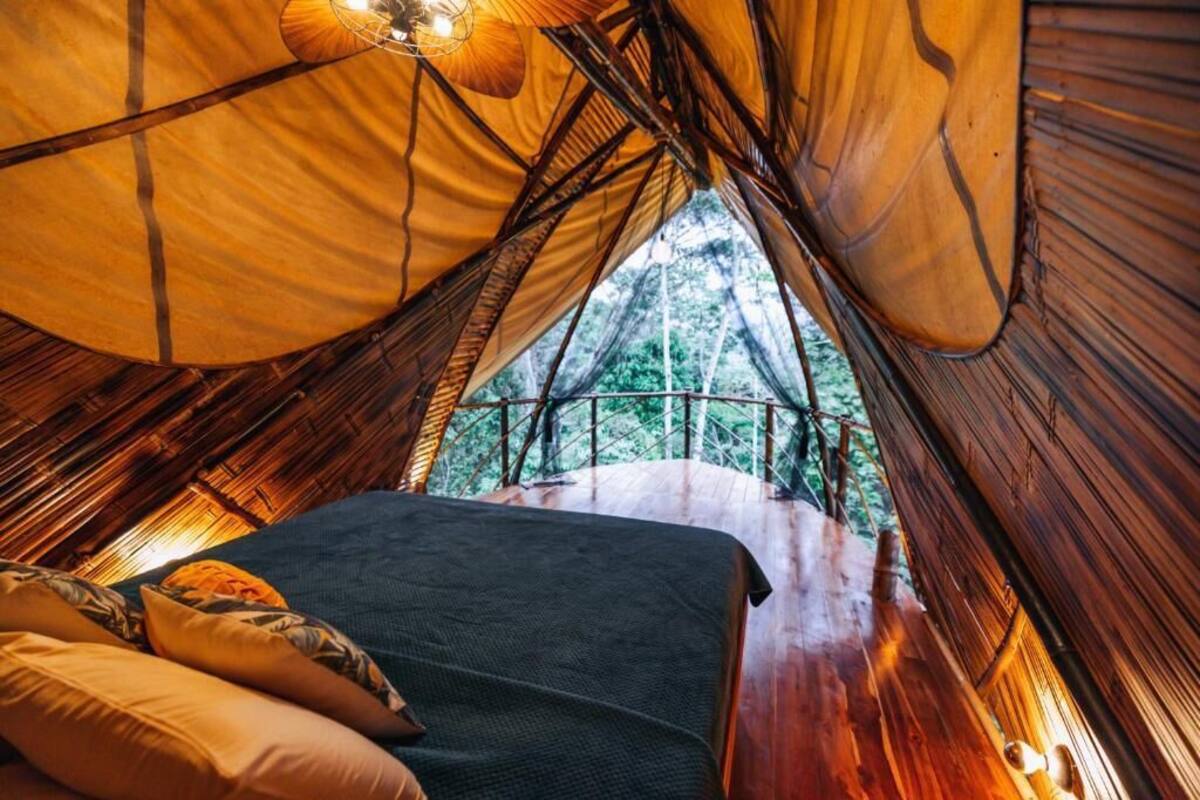 Uma tenda com cama, luminária pendente e vista para árvores pela entrada.