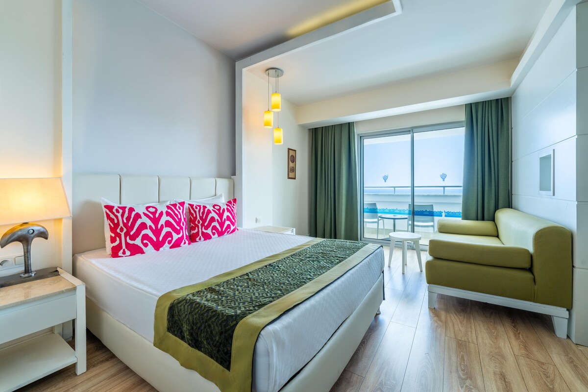 Quarto de hotel com cama, mesa de cabeceira, escrivaninha, poltrona verde e varanda com vista.