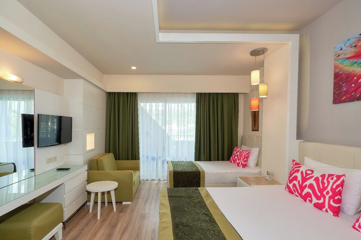 Quarto de hotel com cama, escrivaninha, cadeira, sofá, mesinha e televisão.