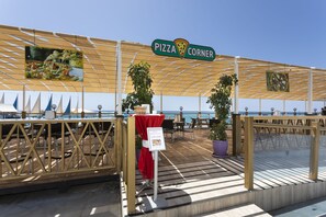 Uma área de refeições ao ar livre coberta, com um letreiro que diz 'PIZZA CORNER', mesas, cadeiras e plantas em vasos.