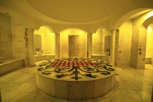 Uma fonte de mármore circular com um tecido quadriculado no centro, rodeada por arcos e colunas em um espaço interior bem iluminado.