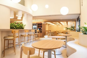 Interior de café moderno com mesas redondas e cadeiras de madeira, balcão de petiscos à vista e plantas em vasos.