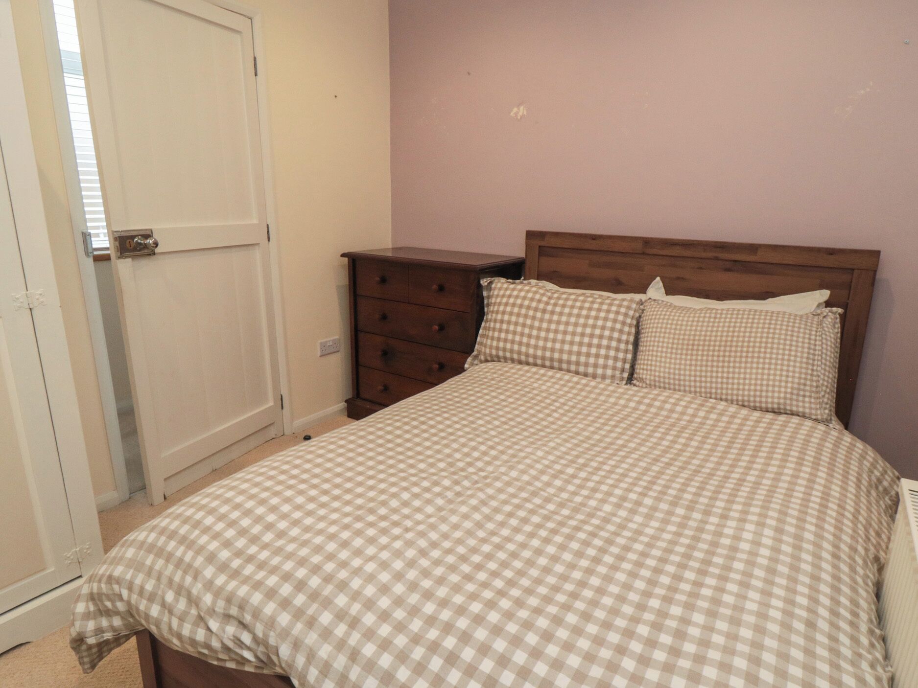 Quarto com colcha xadrez, cómoda de madeira e uma porta.