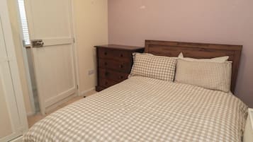 Quarto com colcha xadrez, cómoda de madeira e uma porta.