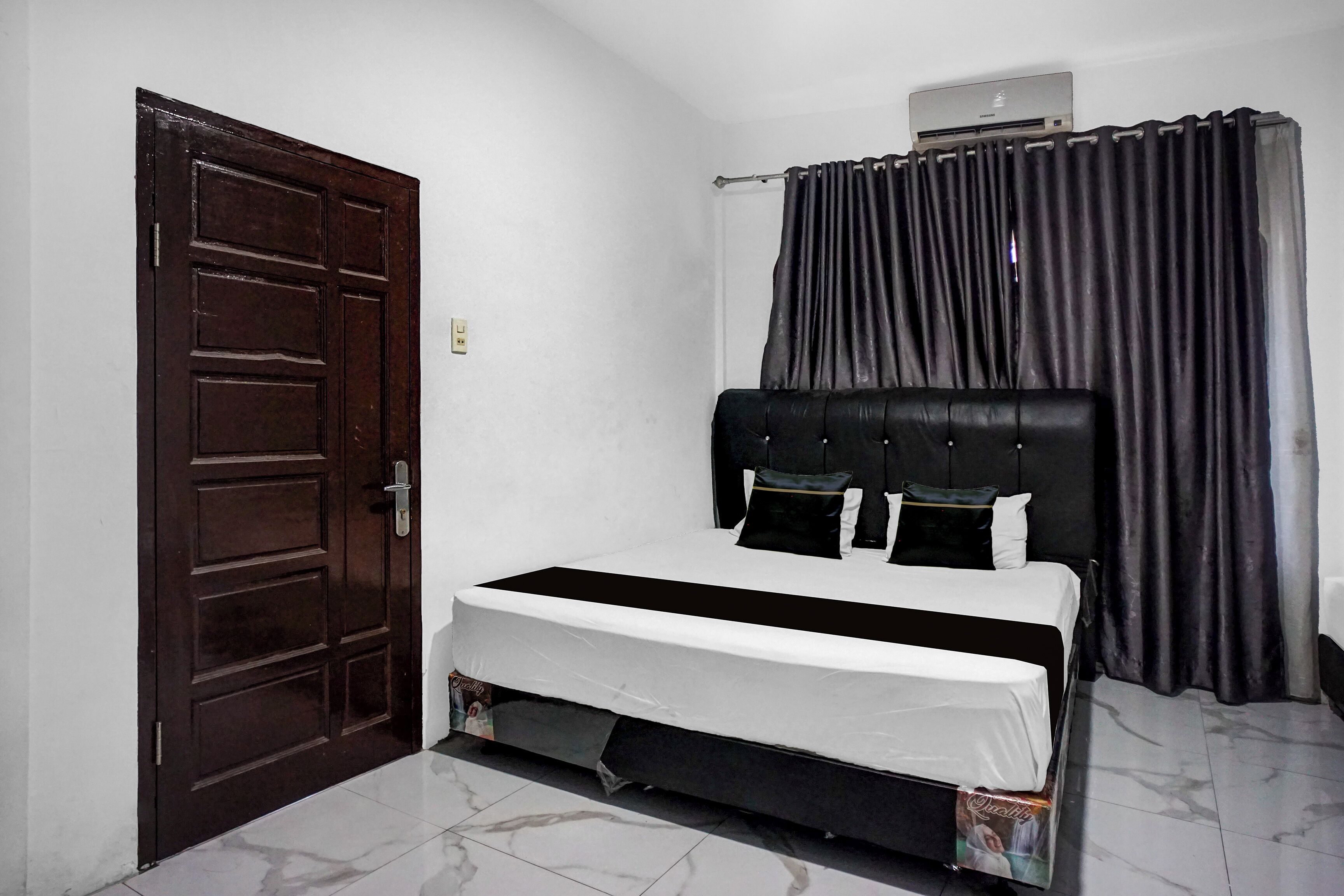 Quarto com uma cama de solteiro, uma porta de madeira escura e uma janela com cortinas pretas.