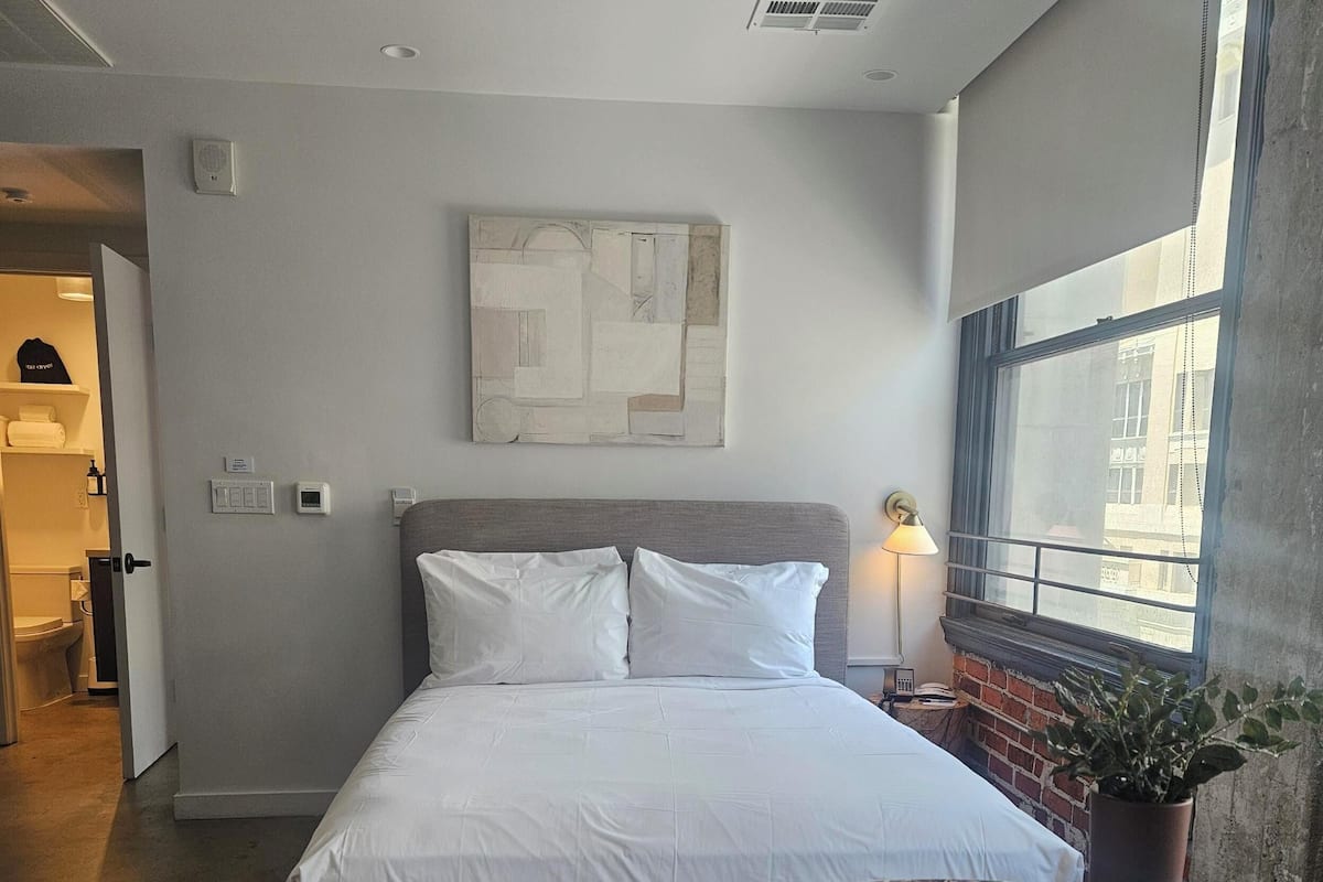 Quarto de hotel moderno com uma cama, uma pequena mesa com um vaso, uma janela com vista para prédios e uma porta que dá para um banheiro.