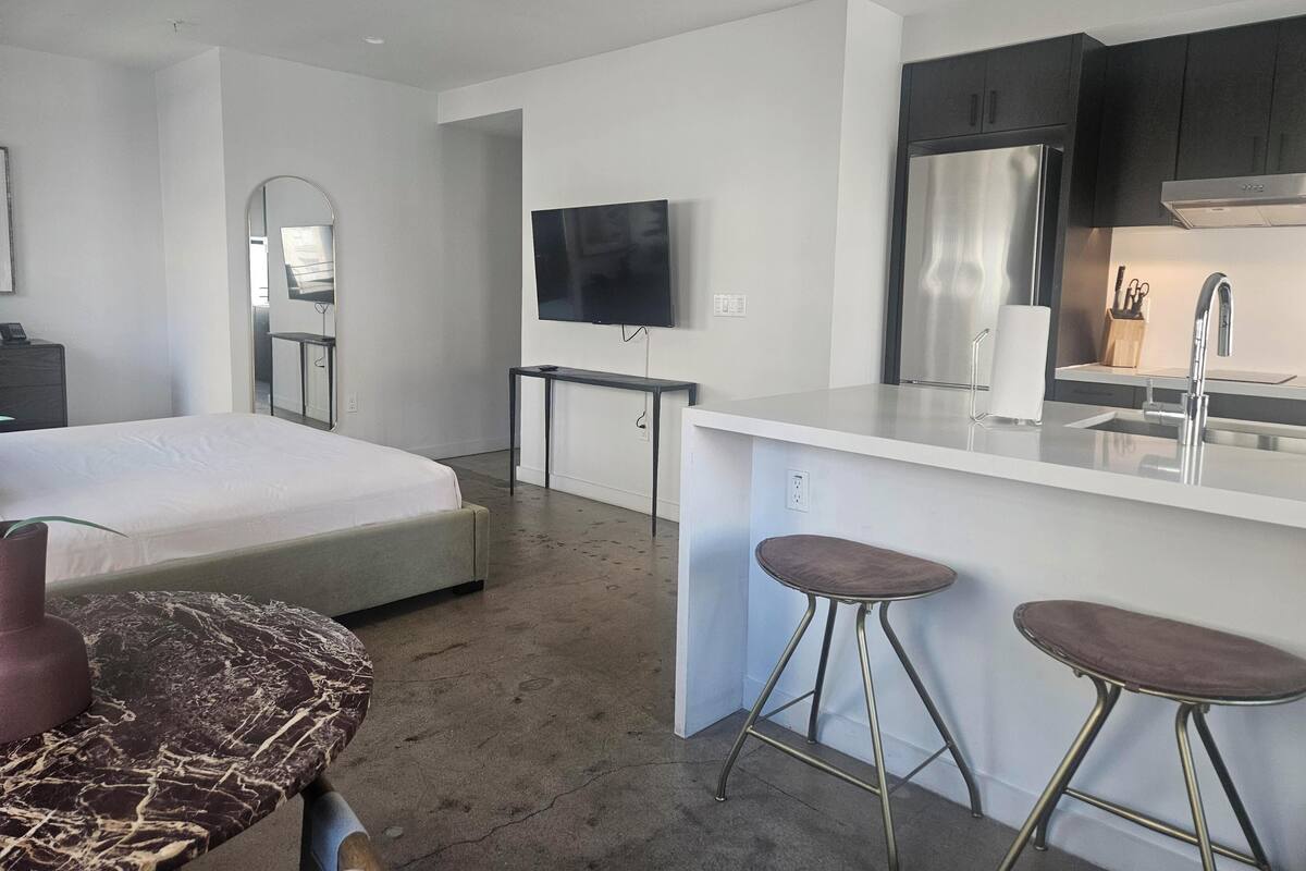 Quarto de hotel moderno com cama, televisão, área de cozinha com frigorífico e um pequeno banco de bar.