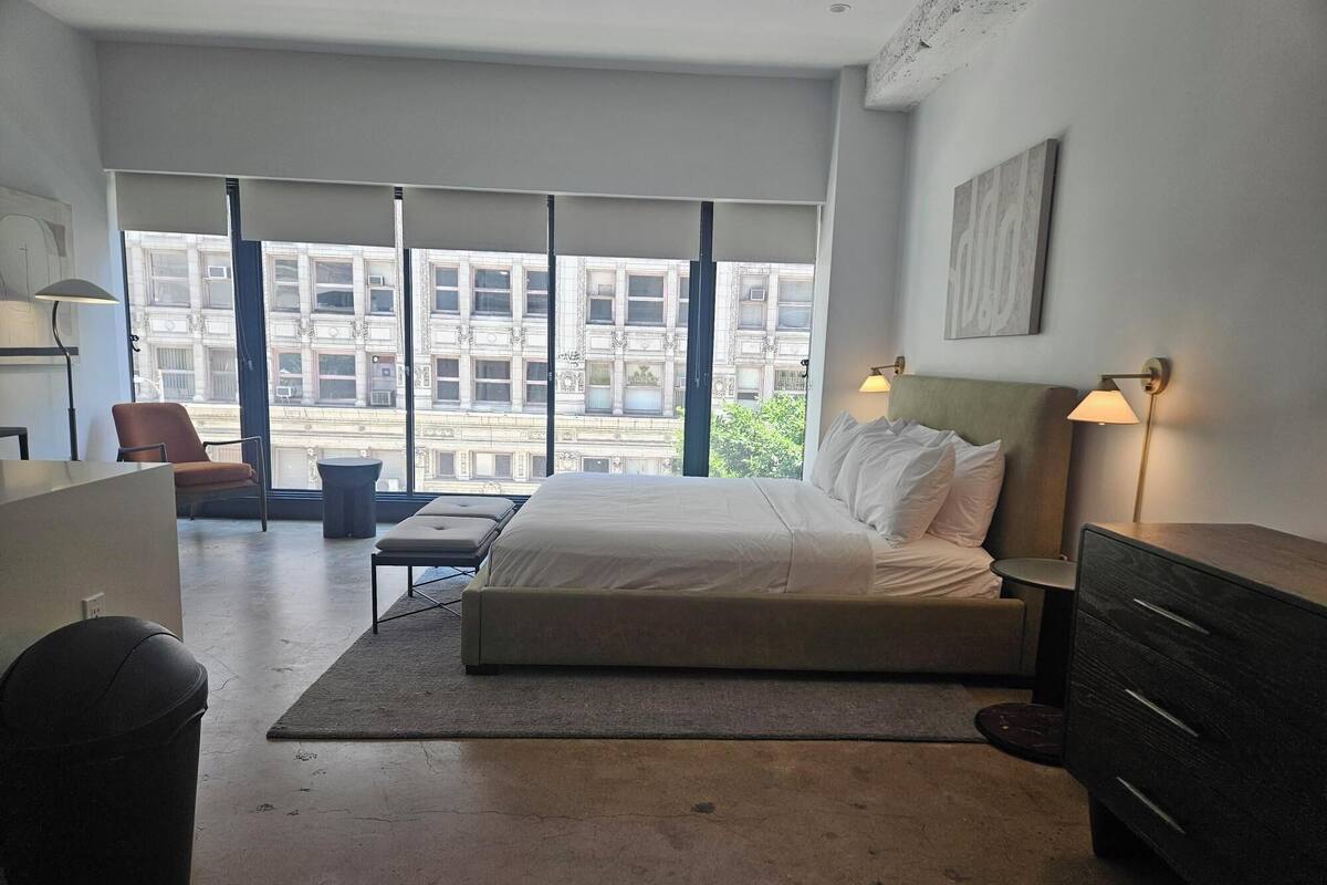 Quarto de hotel moderno com uma cama grande, uma escrivaninha, uma cadeira e vista para um edifício através de amplas janelas.