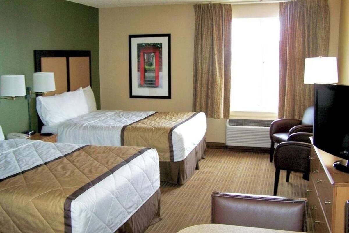 Quarto de hotel com duas camas, uma escrivaninha com cadeira, televisão e um quadro na parede.