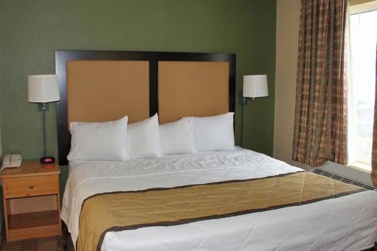 Quarto de hotel com cama, abajures, telefone, janela com cortinas e radiador.